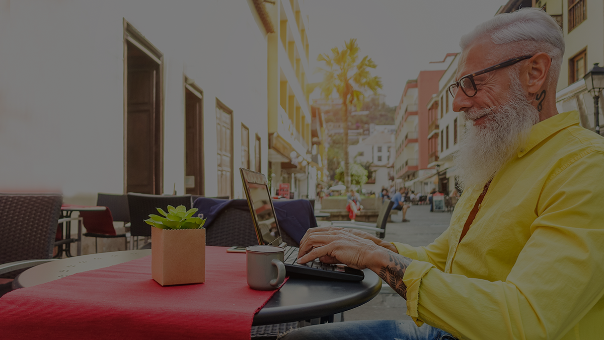 A senior man with a beard and glasses working on a laptop at an outdoor cafe on a sunny day, with buildings and a palm tree in the background.